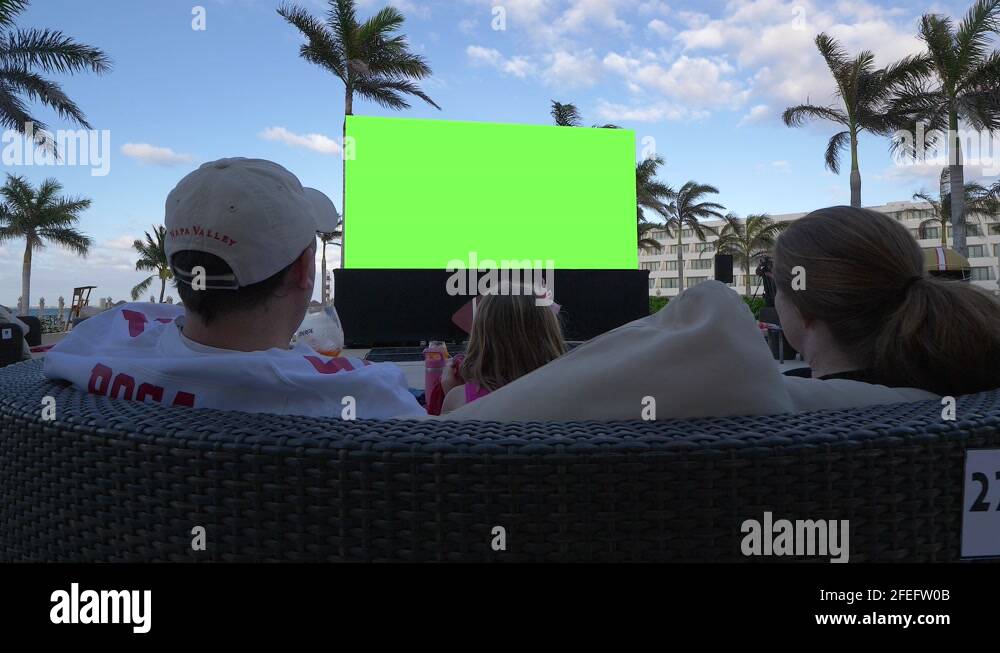 Family of 3 watching large outdoor green screen at a tropical resort ...