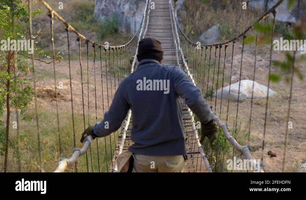 Rear view of man walking on simple suspension bridge over dry sandy ...