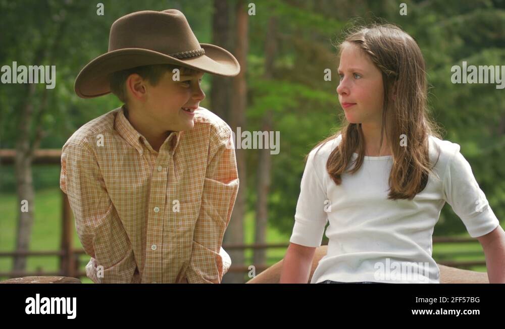 A boy in a cowboy hat sit on a fence with a young girl taking off his ...
