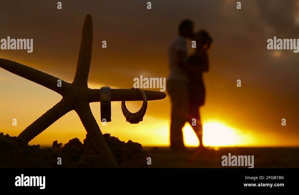 Wedding rings on the starfish and couple kissing at sunset beach love ...