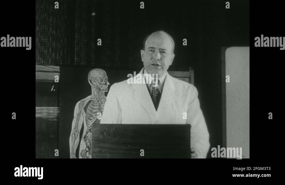 1940s. Doctor delivers lecture from podium. Skeleton chart is behind ...