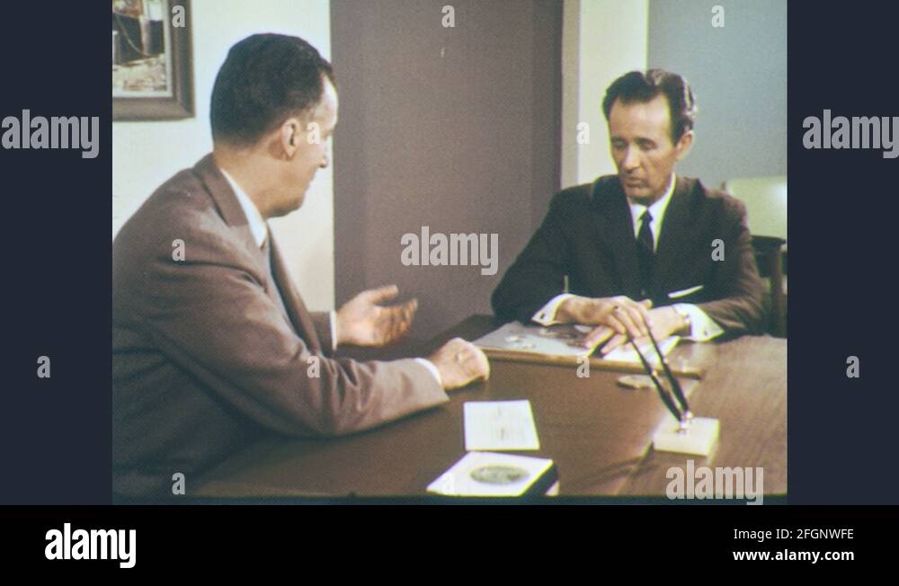 1960s: Two men in suits sit at a desk talking. One man hands the other ...