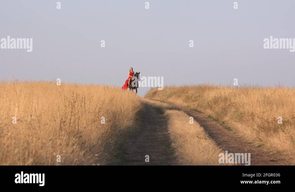 Young girl in long scarlet red dress riding black horse across dry ...
