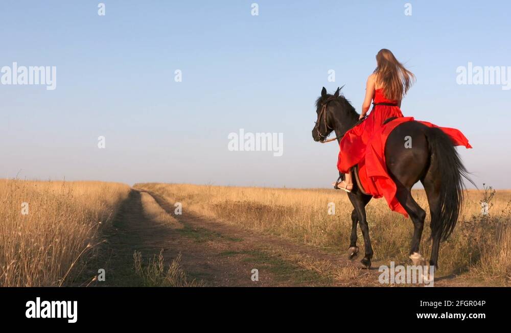 Young girl in long scarlet red dress riding black horse across dry ...