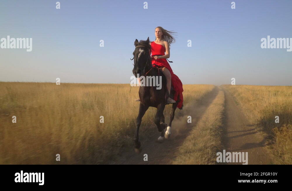 Beautiful girl horseback rider in red dress riding horse across dry ...