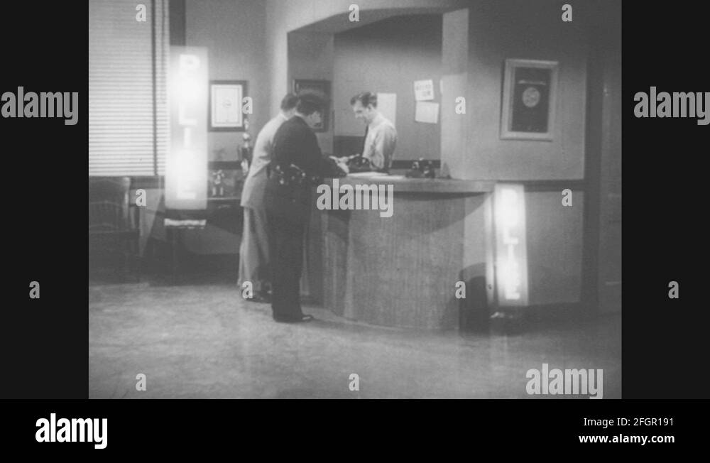 1950s: Policeman in uniform stands at precinct desk. Arrested person ...