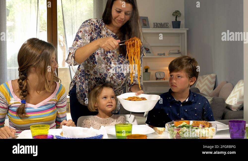 Mother serving spaghetti with tomato sauce to children at the table ...