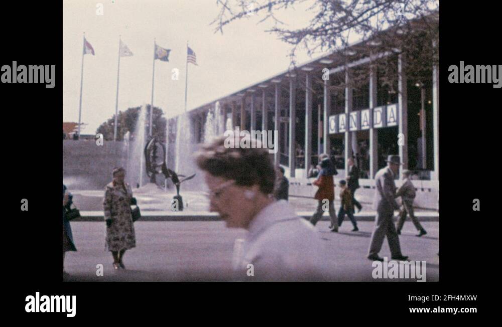 1960s: People walking past fountain. Boy and girl walk past flags ...