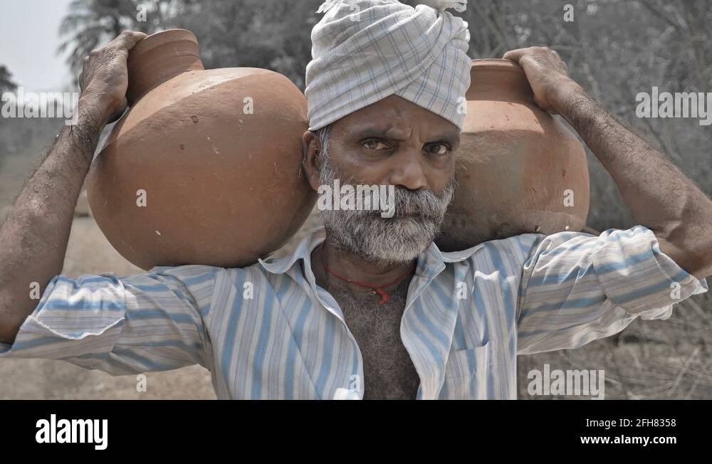 A poor Indian man carrying two empty clay pots standing in dry farmland ...