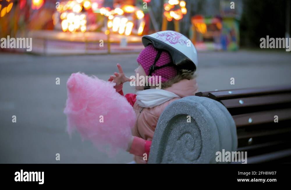 Beautiful little girl sitting on a bench in an amusement park, eating ...