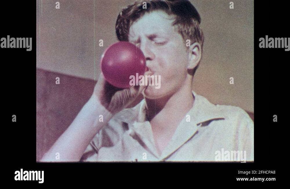 1960s: Boy blows up rubber balloon. Boy pulls balloon neck through hole ...