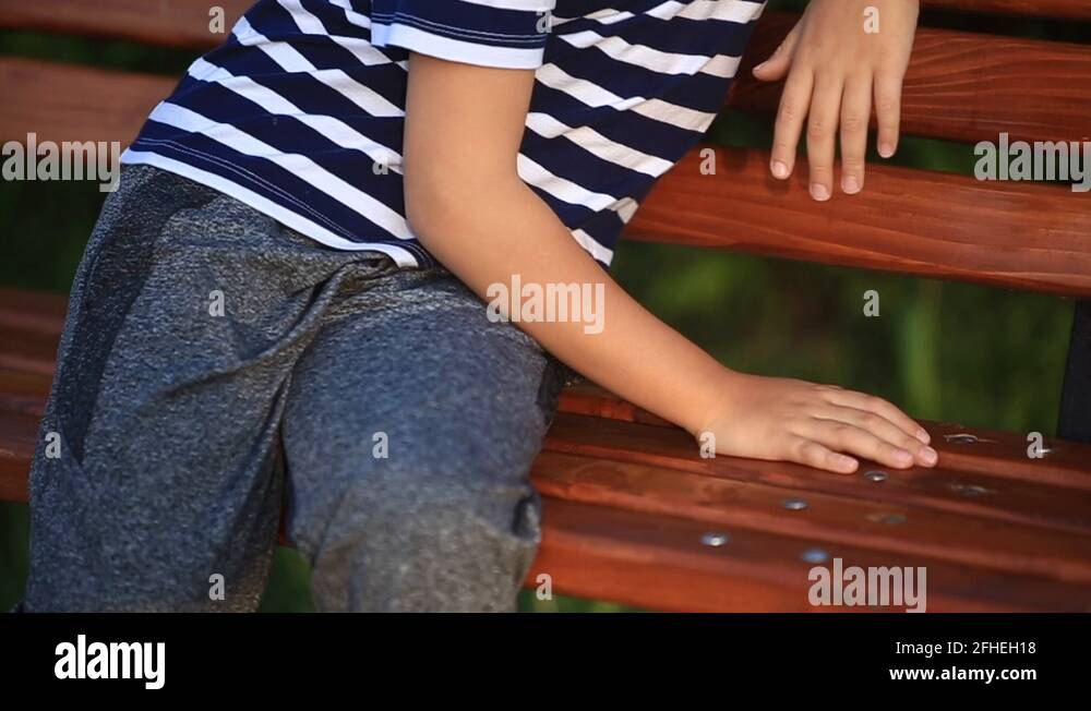 A boy in pants and sneakers sitting on a bench waving his legs. Summer ...
