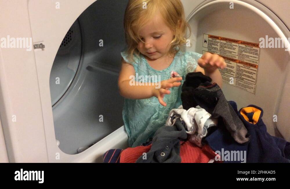 Little girl getting clean clothes out of dryer doing housework with mom ...