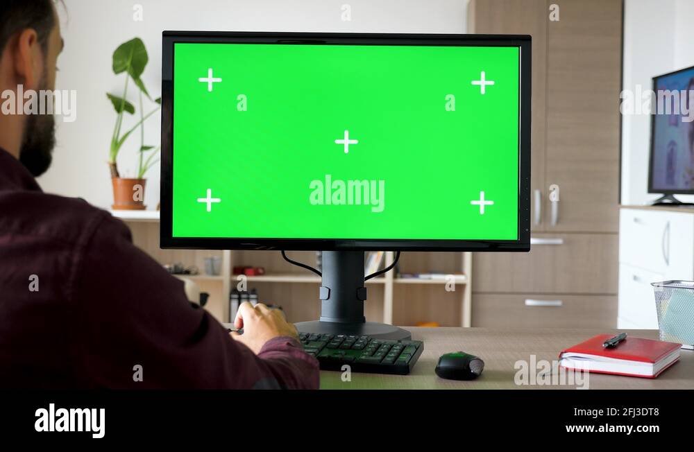 Side view of man sitting in front of computer with big green screen ...