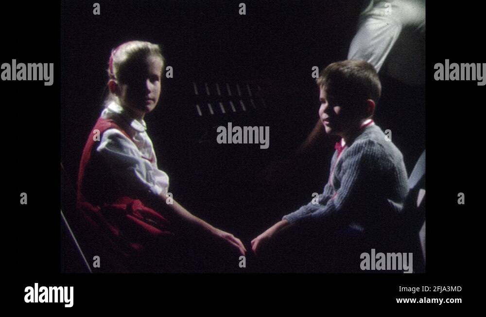 1960s: Boy and girl sit across from each other. Man behind them holds ...