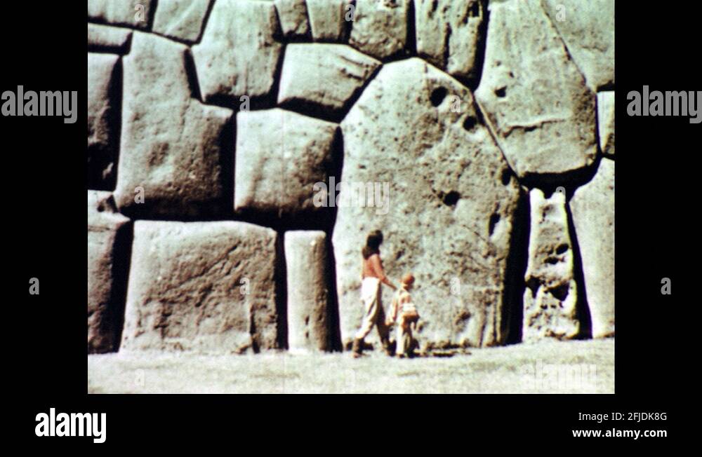 1960s: Woman and child stand next to stone wall. Man walks next to ...