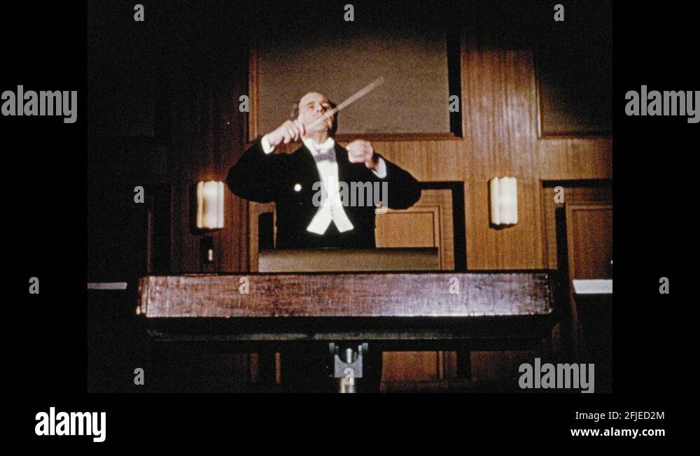 1950s: Man stands conducting orchestra. Two men playing violins in ...