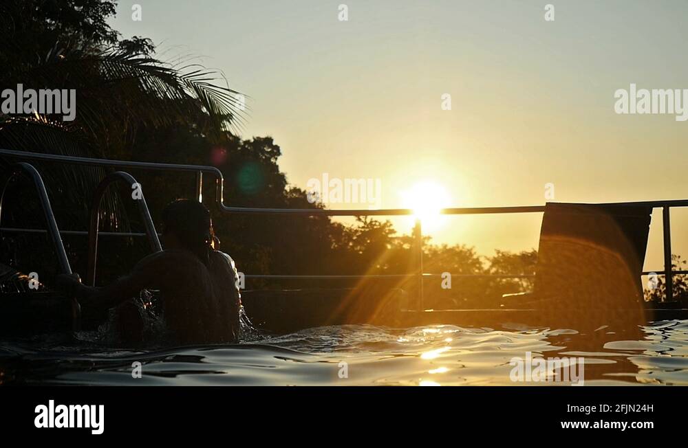 Young man jumps in swimming pool water at Sunset through the sun with ...