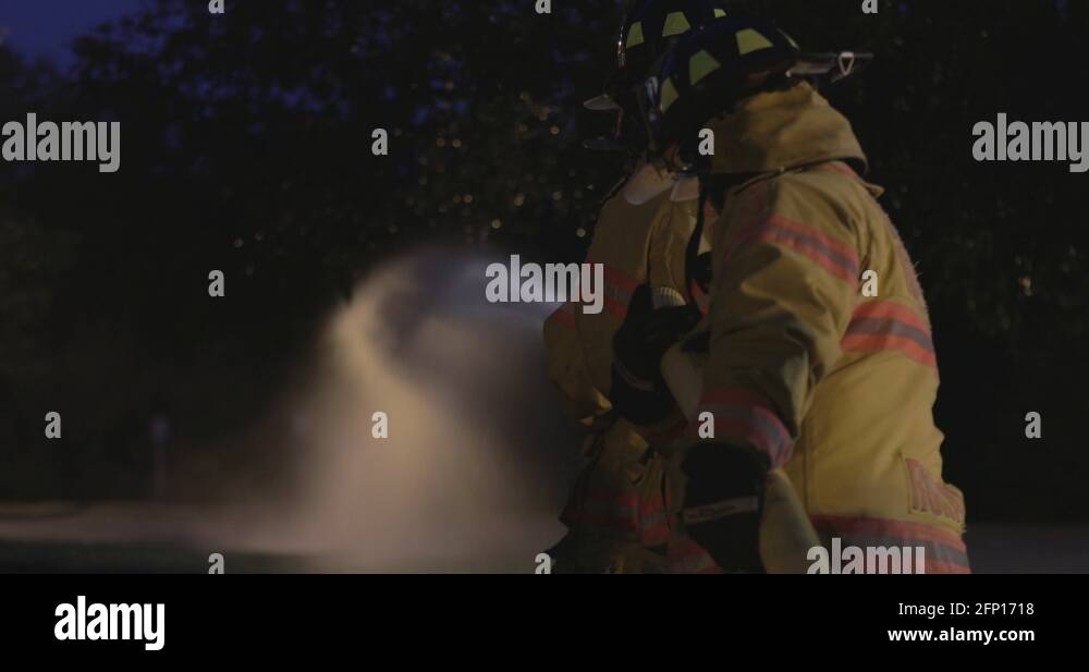 Firefighters spray water with a fire hose near a fire engine at night ...