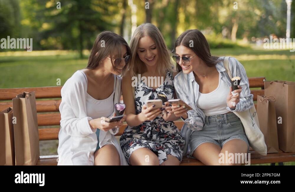 Three attractive girls together eating ice cream cones on a park bench ...