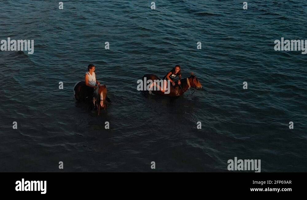 Two girls astride horses in shallow water splash each other, having fun ...