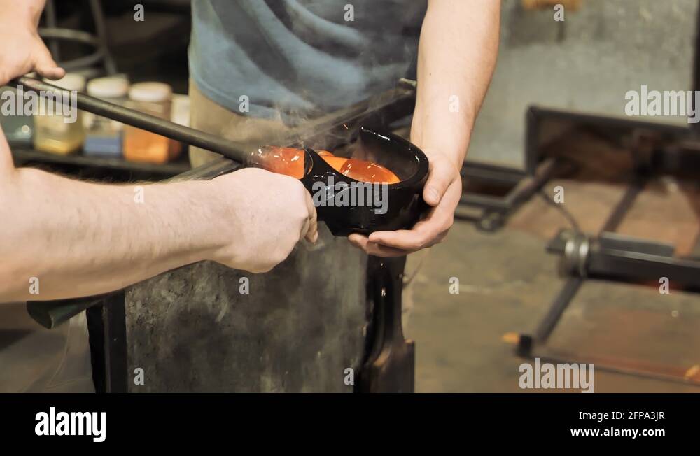 Two glass artists form red hot molten glass by rotating inside a mold ...