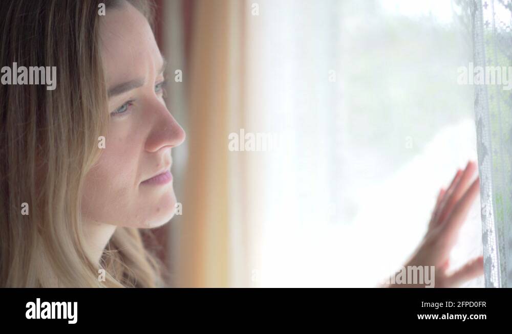 Sad girl inside house looks out through window with white lace curtains ...
