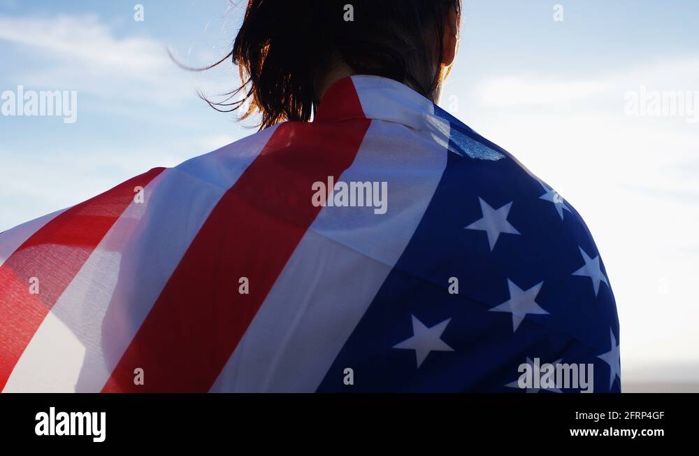 US flag wrapped around an unseen female as she watches the landscape ...
