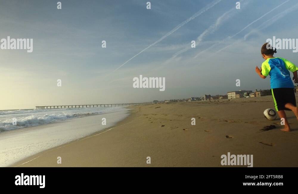 A boy kicks a soccer ball on the beach at sunset with the ocean and ...