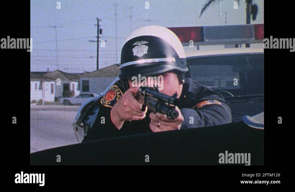 1970s: Police officer kneels behind car, points gun toward camera ...
