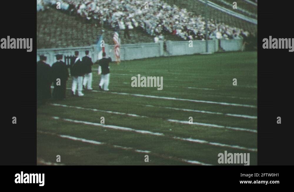 1950s: men and women in graduation gowns march onto football field ...