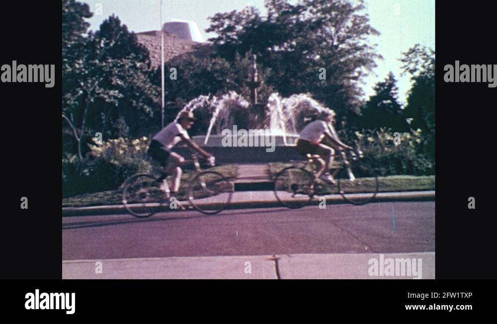 1970s: Two men ride bikes past fountain. Man rides bike on street. Man ...
