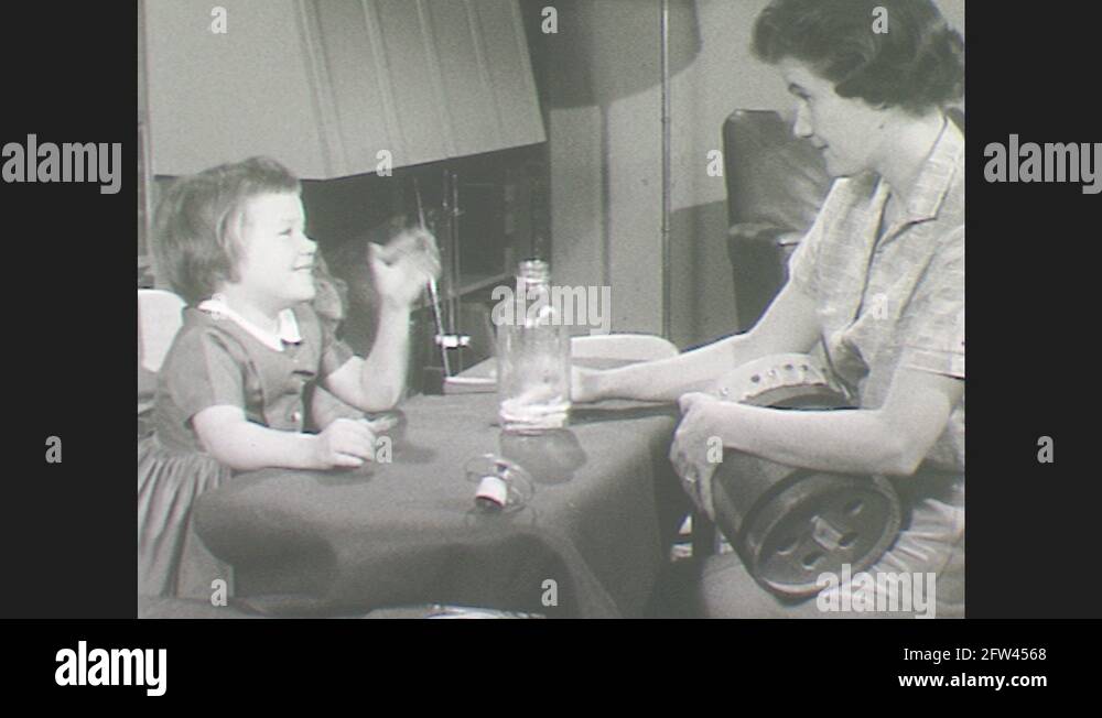 1960s: Woman and girl sit at table. Woman hits drum, girl drops clothes ...