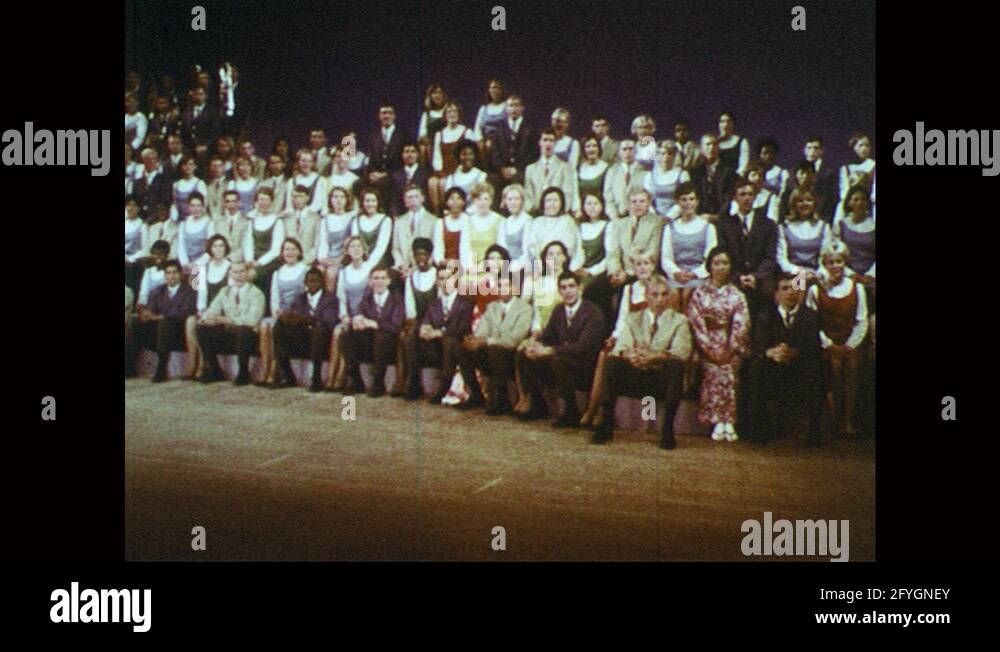 1960s: Choir seated on stage, singing. Close up of man talking Stock ...