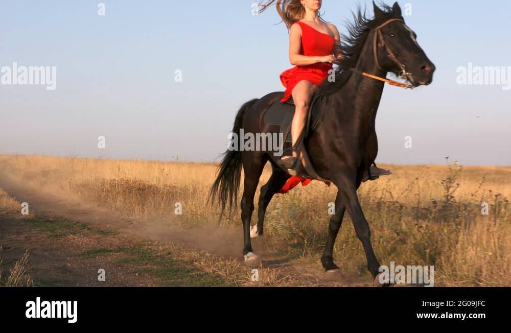 Young girl in long red dress riding black horse on path across dry ...