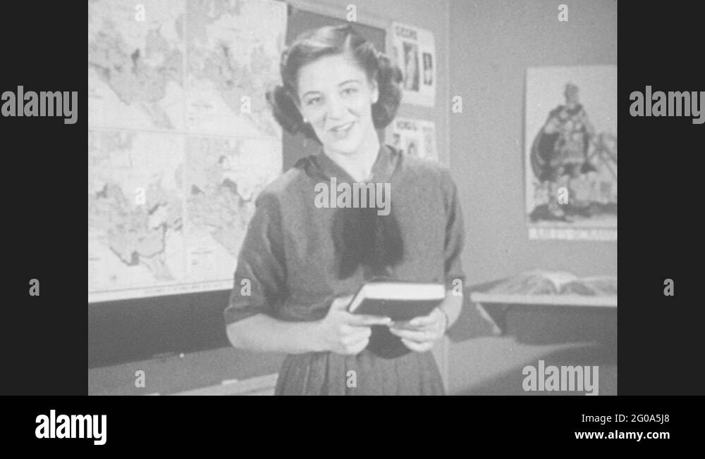 1950s: Teacher in front of classroom talks while holding book. She sets ...