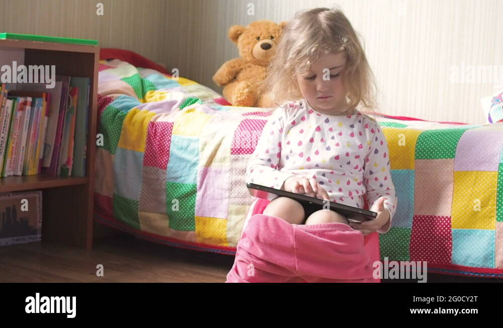 Little girl sitting on the potty beside the bed and playing on a tablet ...