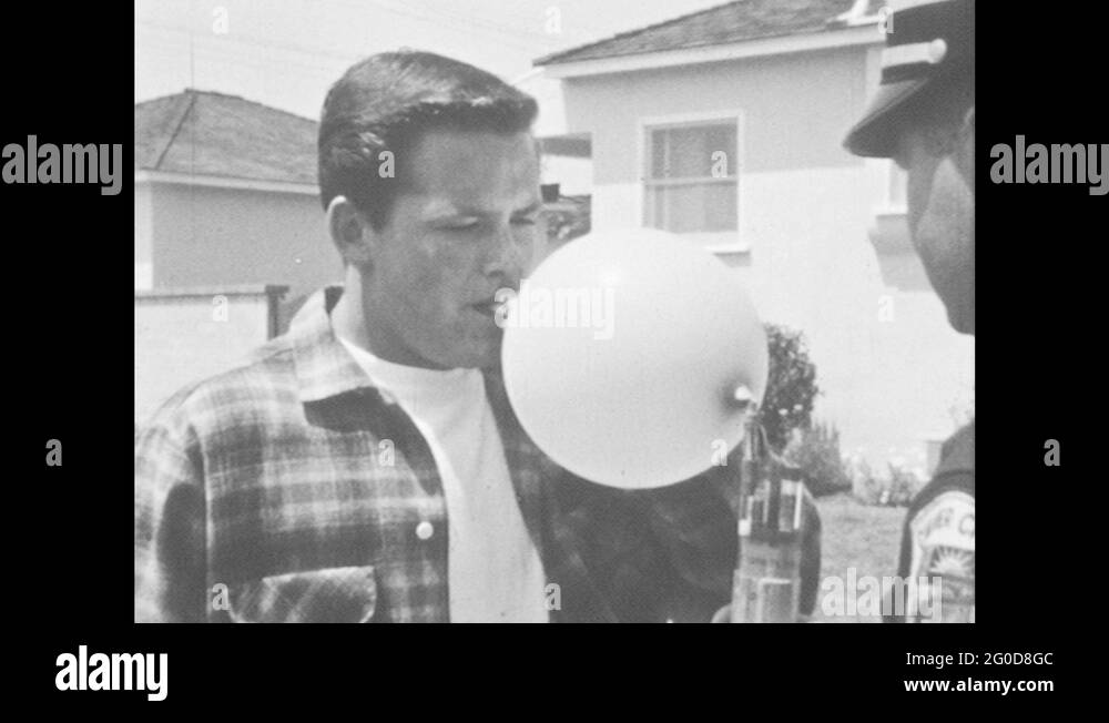 1960s: Young man inflates balloon for DUI breath test. Bottle of ethyl ...