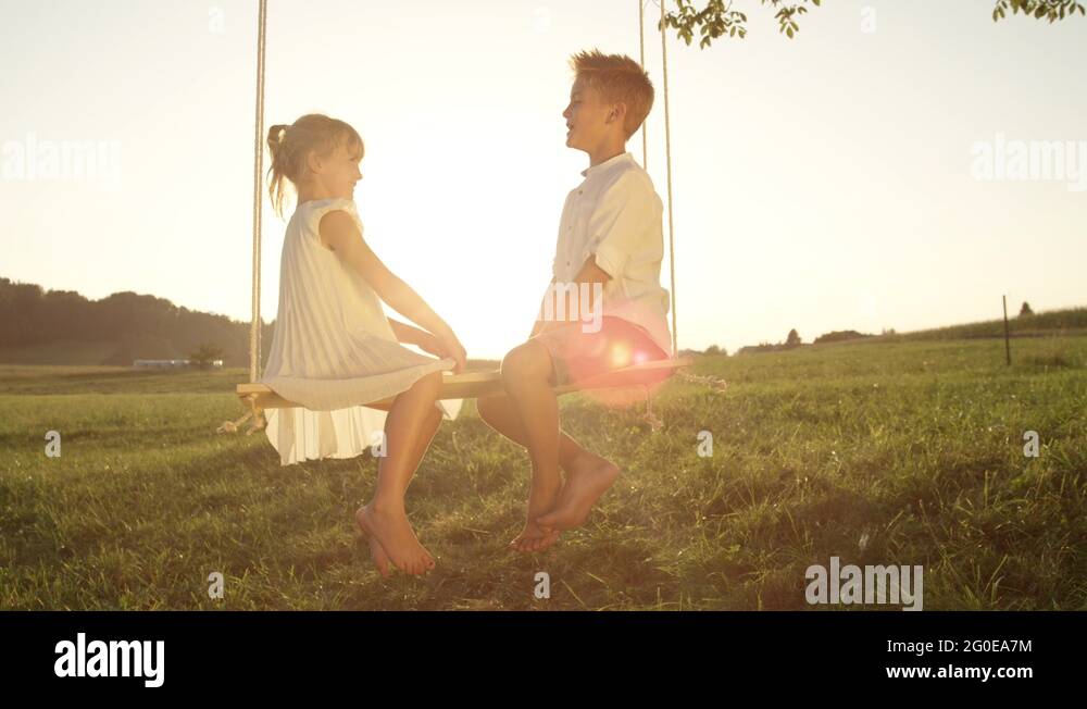 SLOW MOTION Happy children sitting on swing facing each other on warm ...