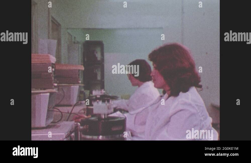 1970s: Two workers sit at table with lab equipment. Man reaches in lab ...