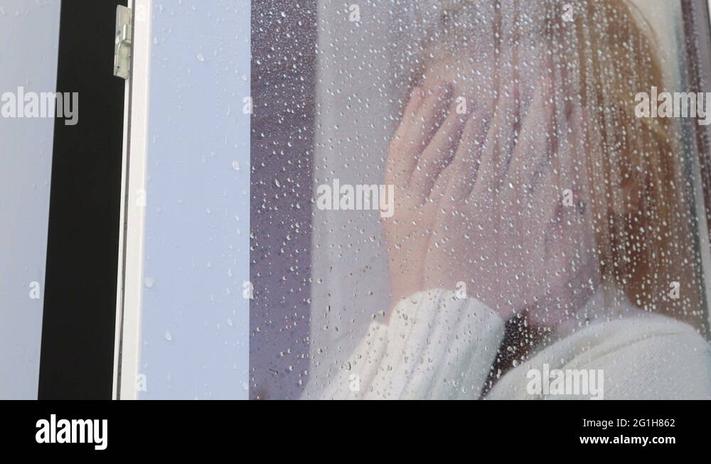 Sad girl closing face with his hands behind window glass with raindrops ...