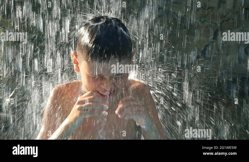 little boy stands under the spray of water of the waterfall , slow ...