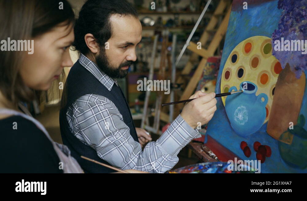 Skilled artist man teaching young girl to draw paintings and explaining ...