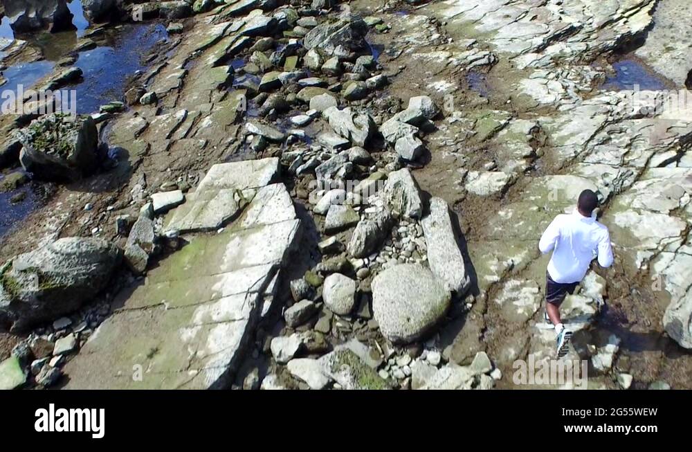Aerial birds eye view shot of a young man running on a rocky ocean ...