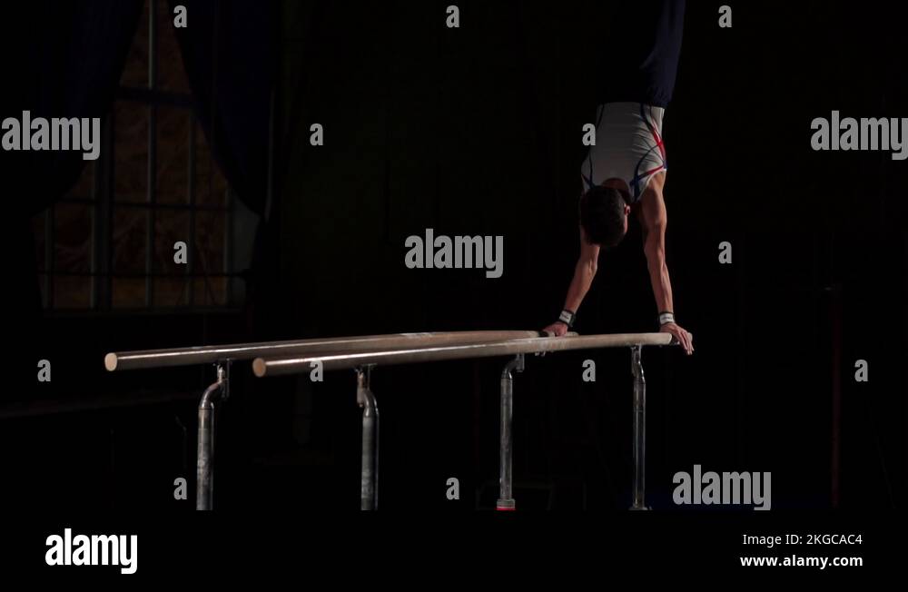 Male gymnast acrobat performs handstand on parallel bars in a dark room ...