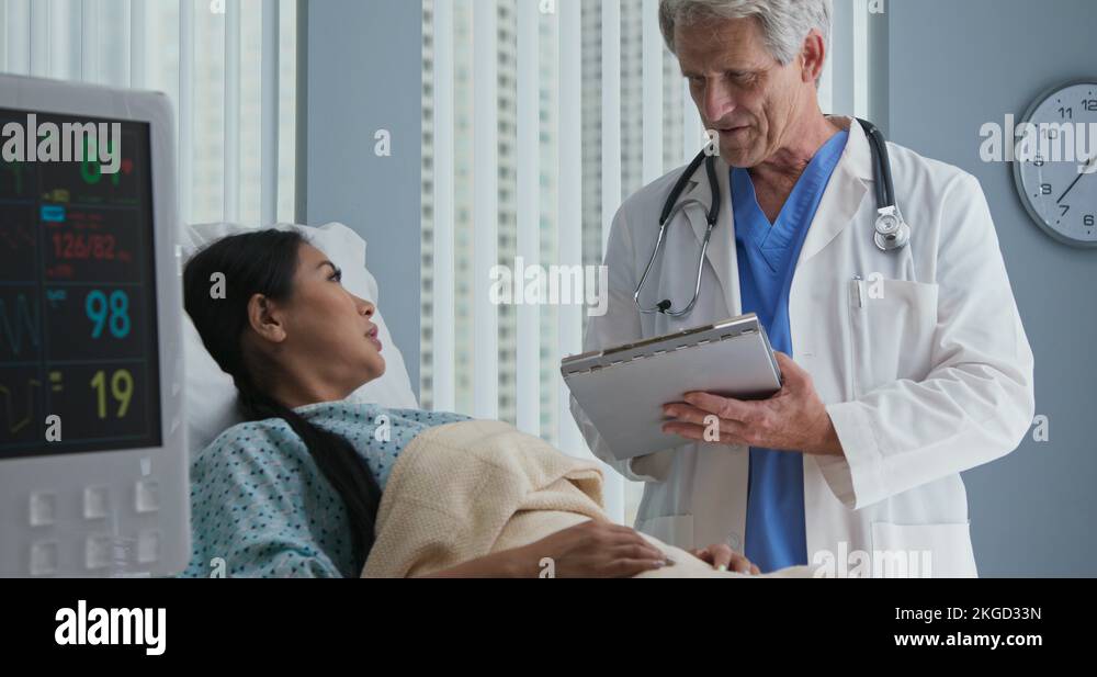 Older male doctor going over chart with female patient lying in ...