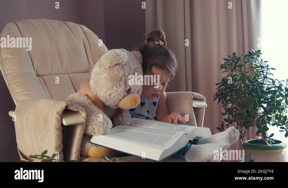 Little girl sitting on the chair reading a book hugging toy teddy bear ...
