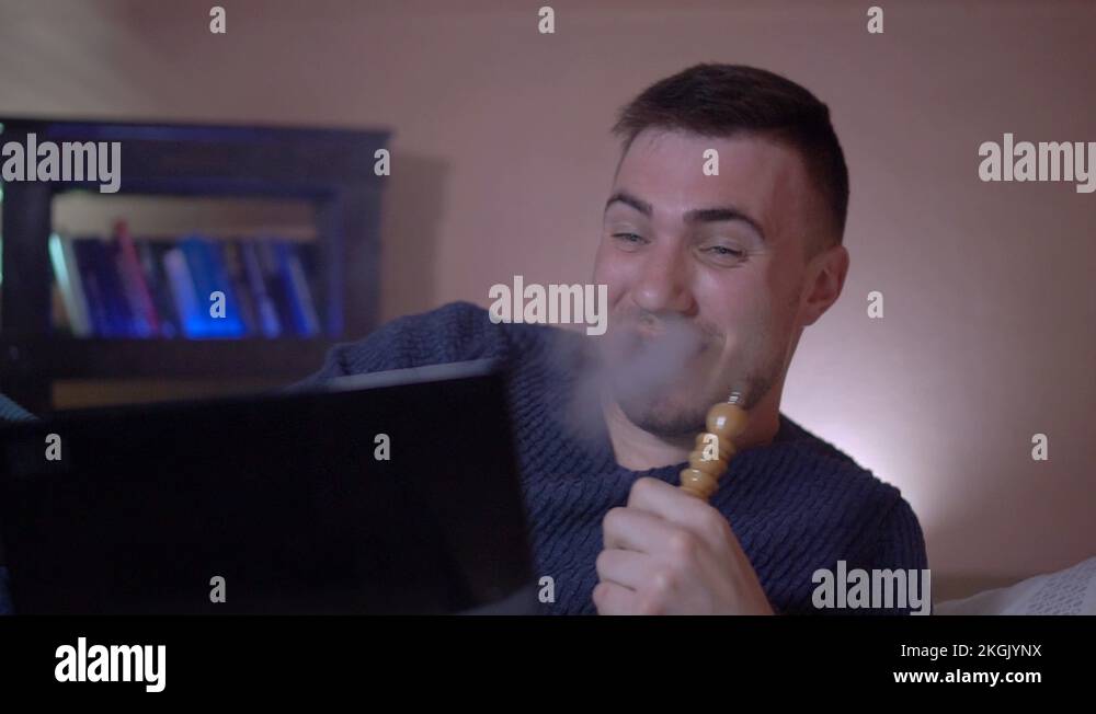 A young guy smokes a hookah, exhales smoke and choked smoke, watching a ...
