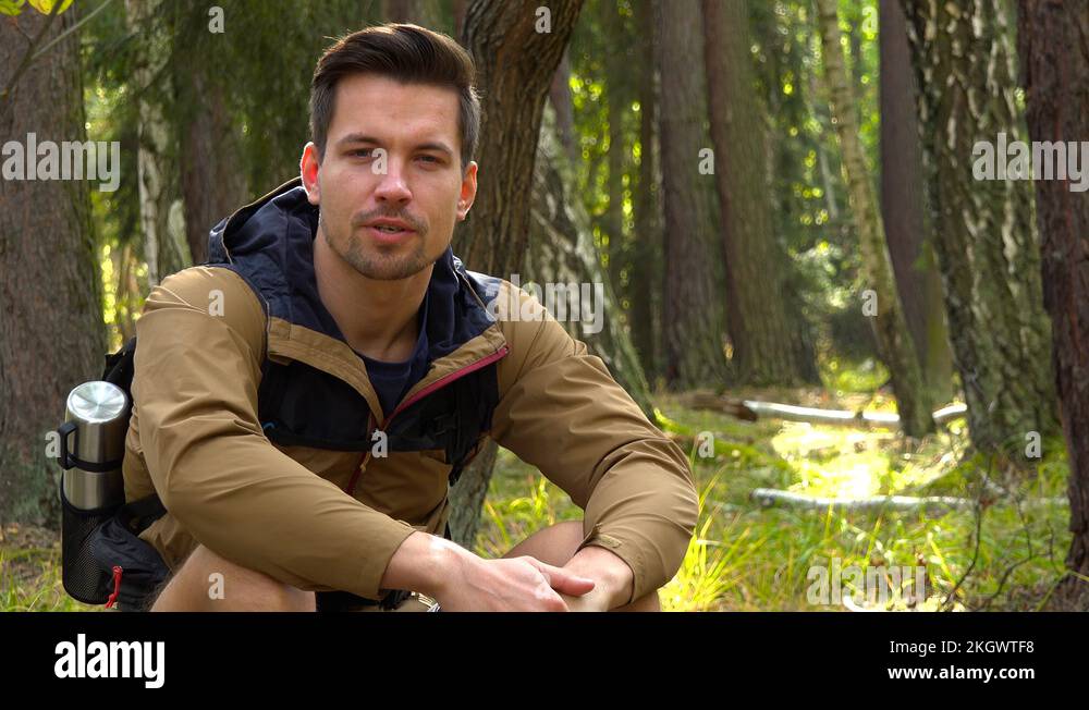 A young handsome hiker sits on the ground in a forest and talks to the ...