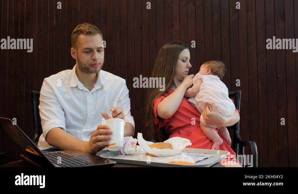 Young parents with baby child mother and father sit eating burger and ...